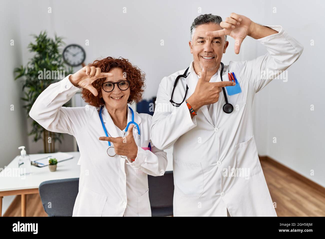 Two middle age doctors at medical clinic smiling making frame with ...