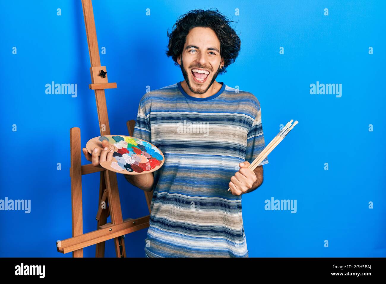 Handsome hispanic man standing drawing with palette by painter easel ...
