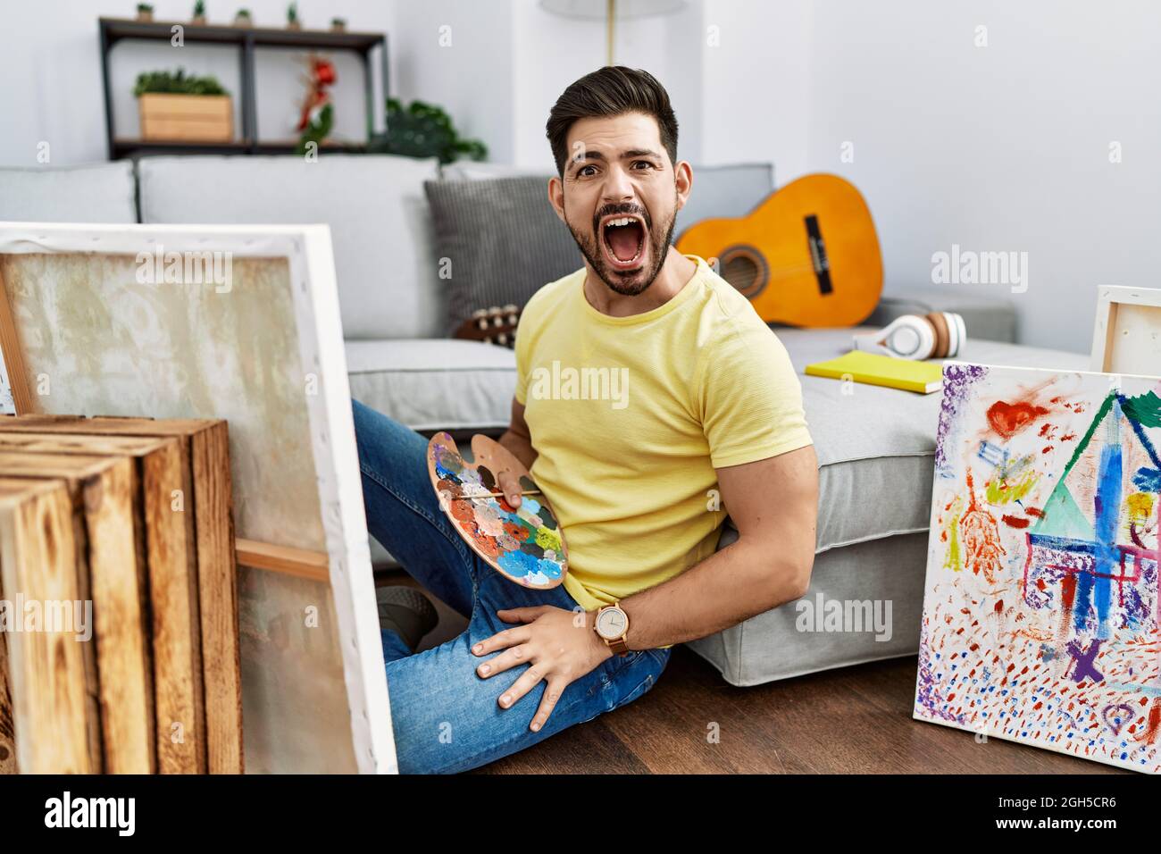 Young man with beard painting canvas at home angry and mad screaming ...