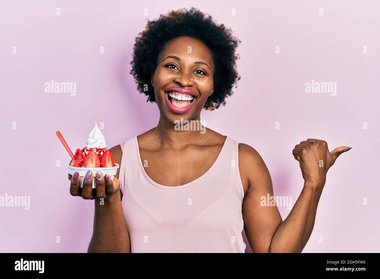 Young african american woman eating strawberry ice cream pointing thumb ...