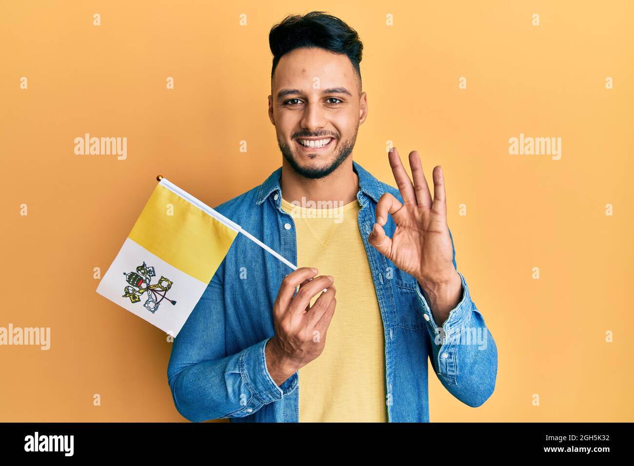 Young arab man holding vatican city flag doing ok sign with fingers ...