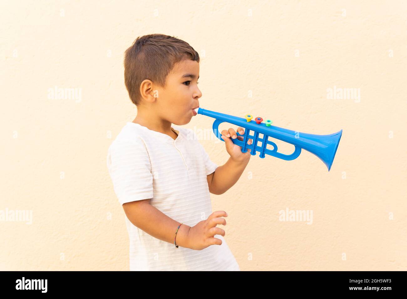 Side view of cute little boy playing blue toy trumpet standing near ...
