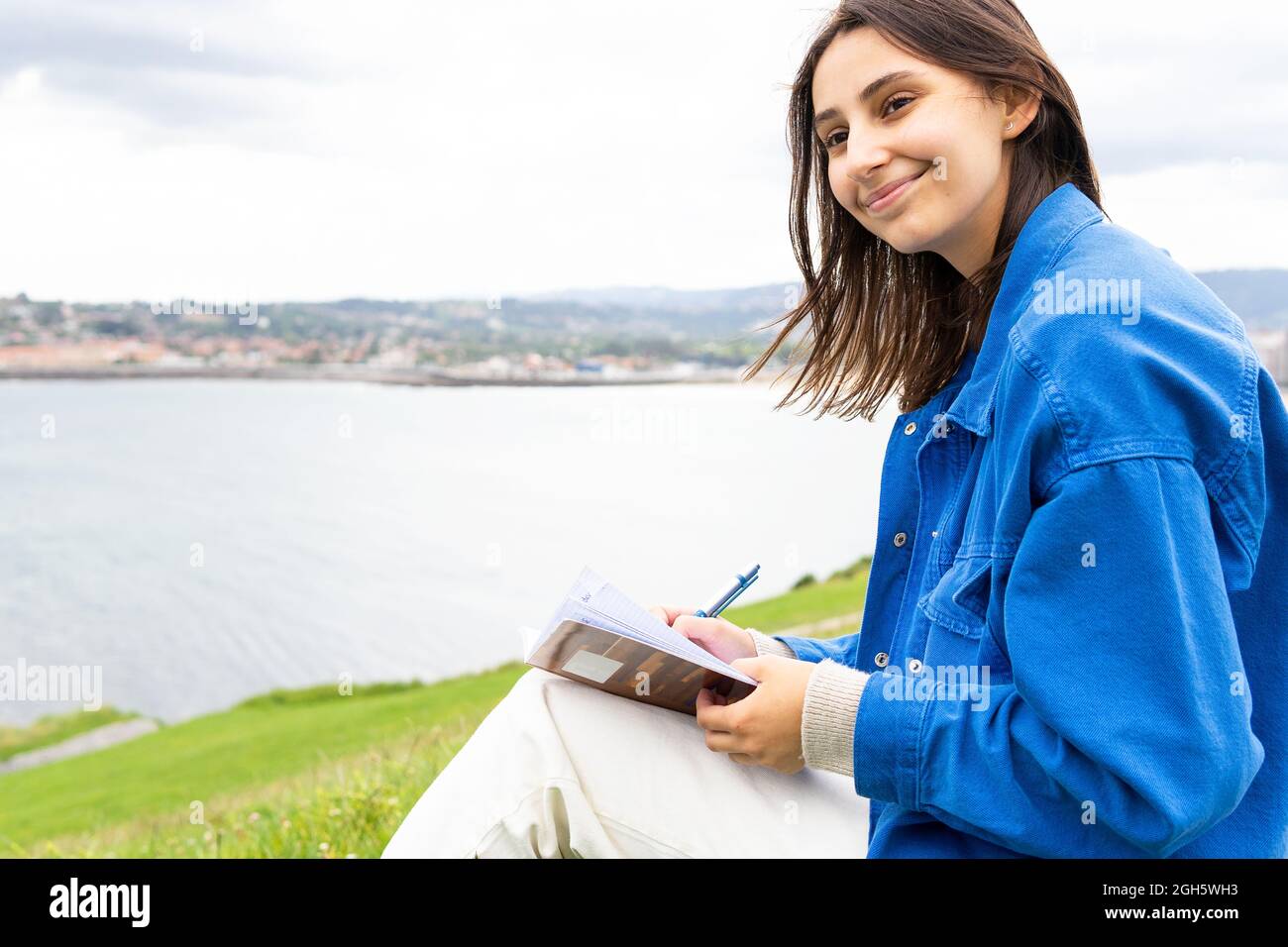 Happy female writing in notepad while sitting on hill near sea and ...