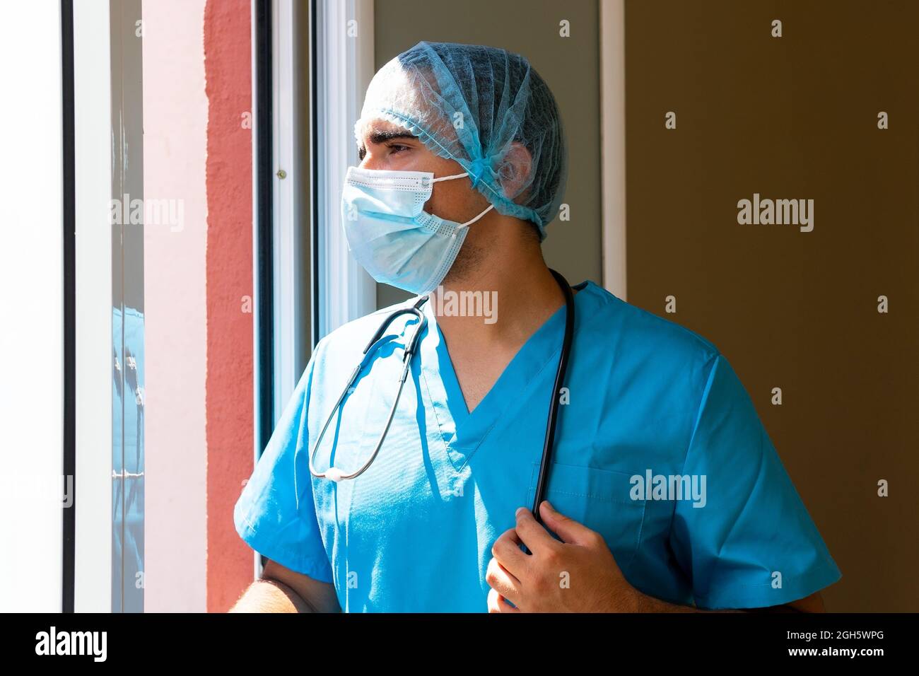 Side view of male medic in mask and uniform standing near window in ...