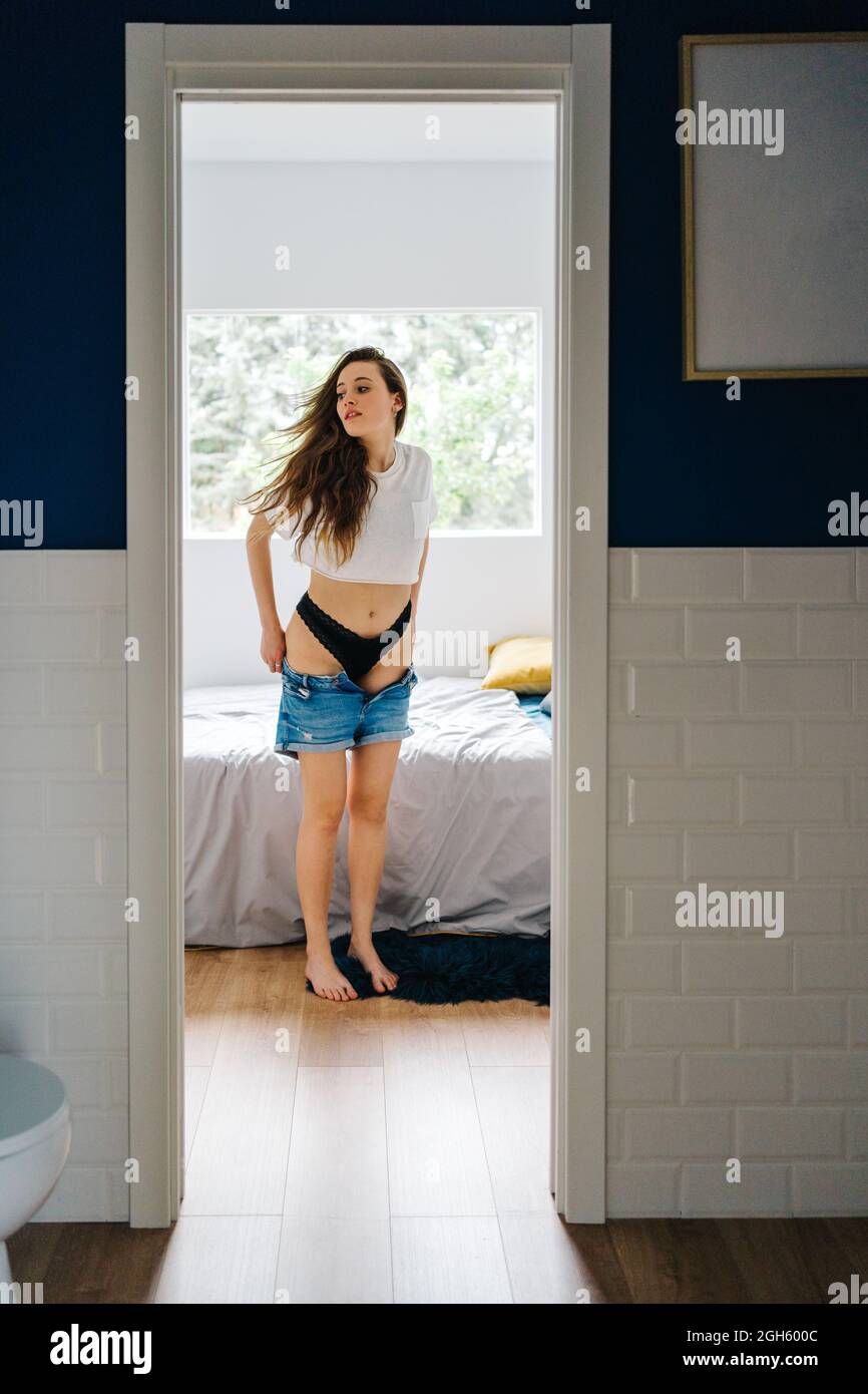 Charming slim female putting on denim shorts while standing in bedroom ...