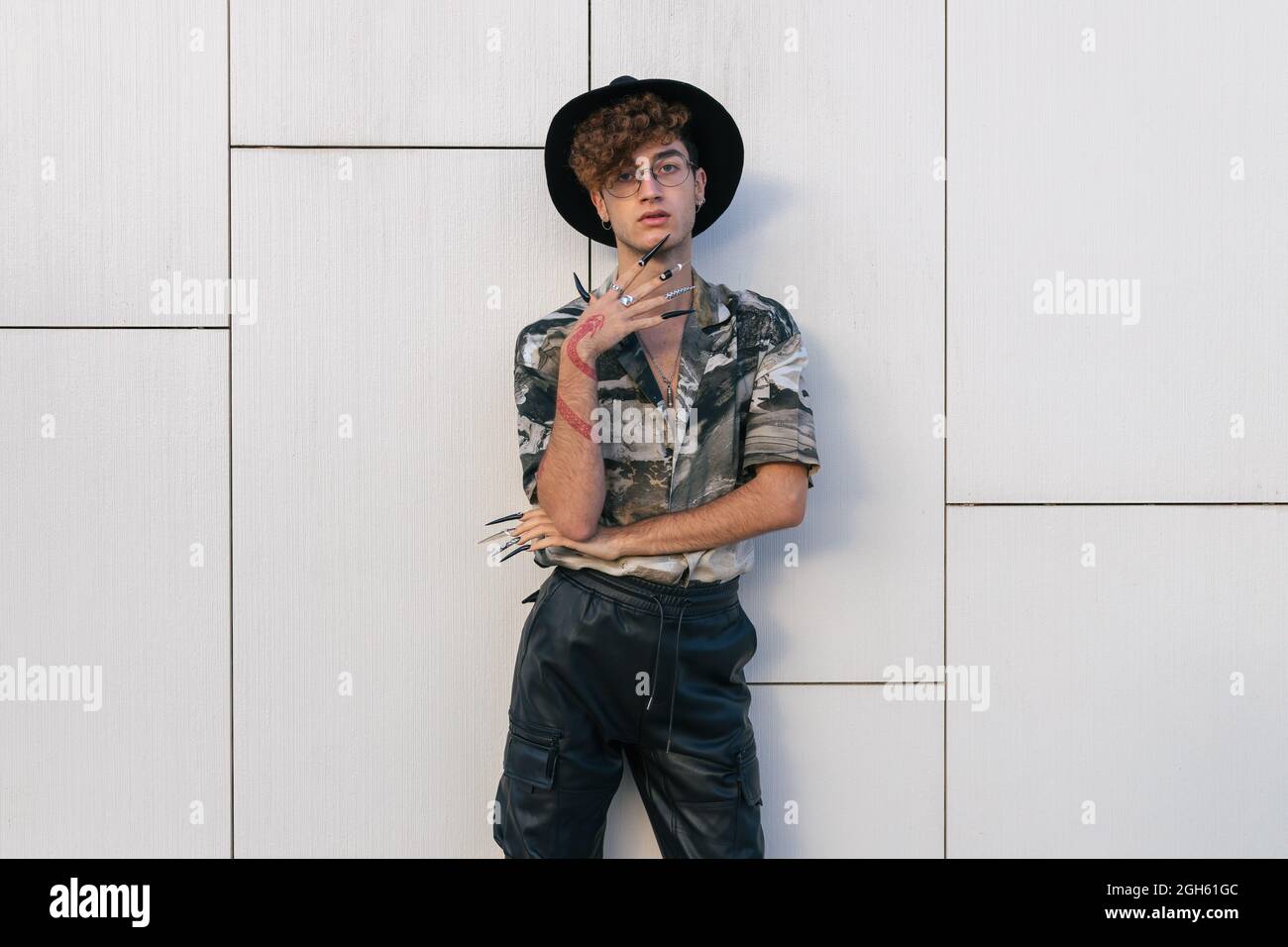 Young vain man in stylish wear with hat standing on tiled wall looking ...