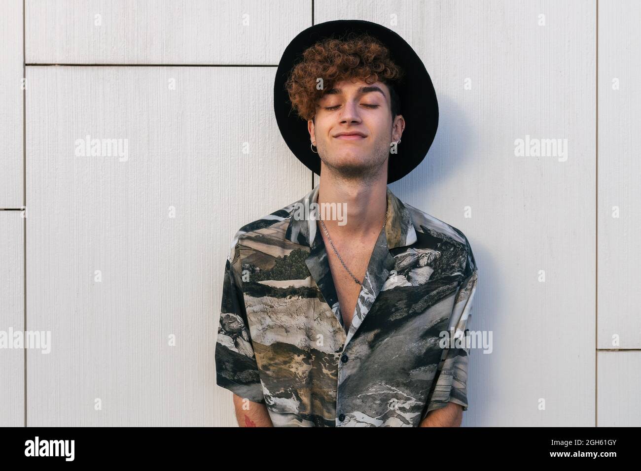 Young vain man in stylish wear with hat standing on tiled wall looking ...