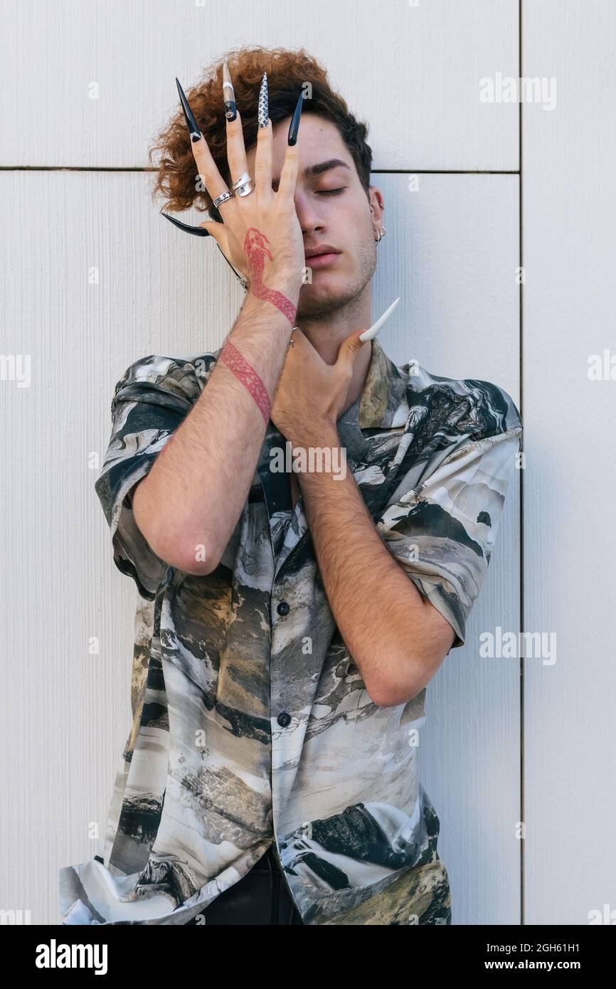 Young vain man in stylish wear with long nails standing on tiled wall ...