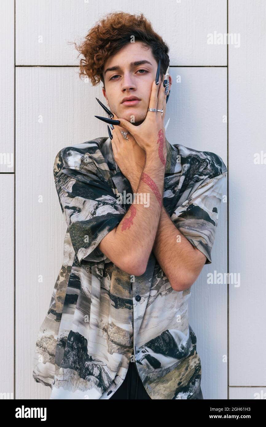 Young vain man in stylish wear with long nails standing on tiled wall ...
