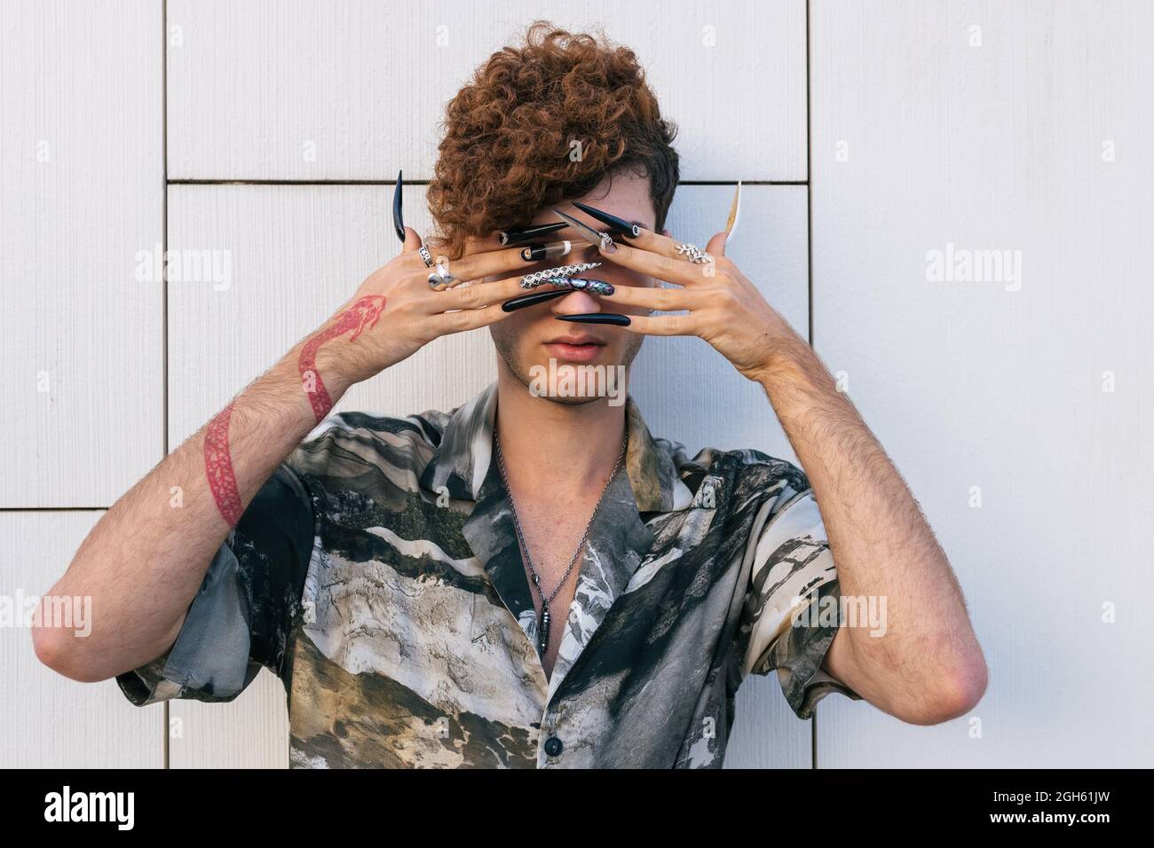 Young vain man in stylish wear with long nails standing on tiled wall ...
