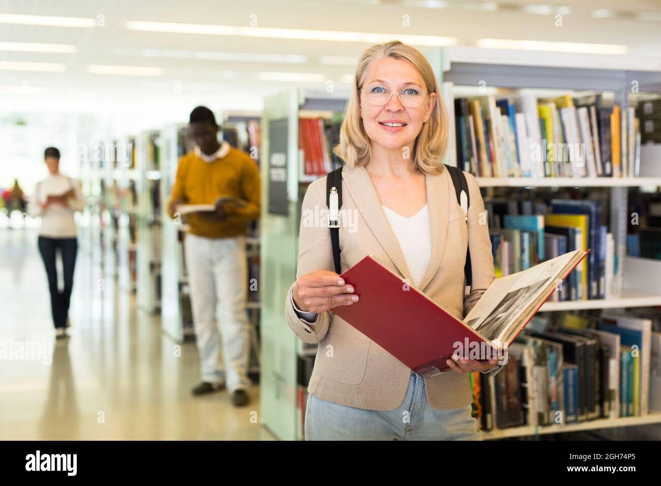 Mature woman reading book while standing near bookshelves in public ...