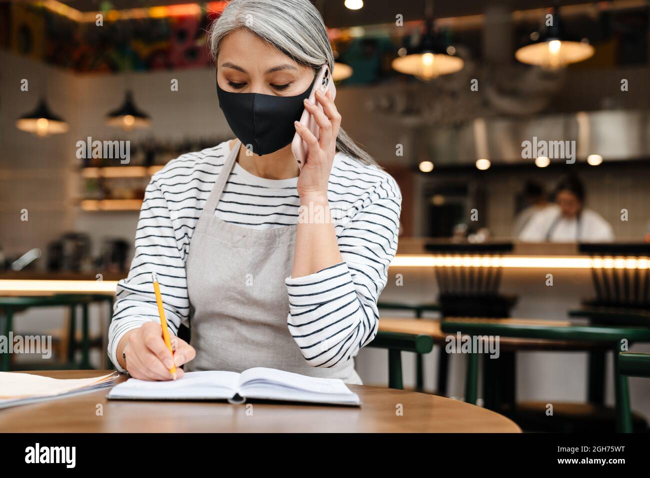 Mature waitress woman wearing face mask talking on cellphone while ...