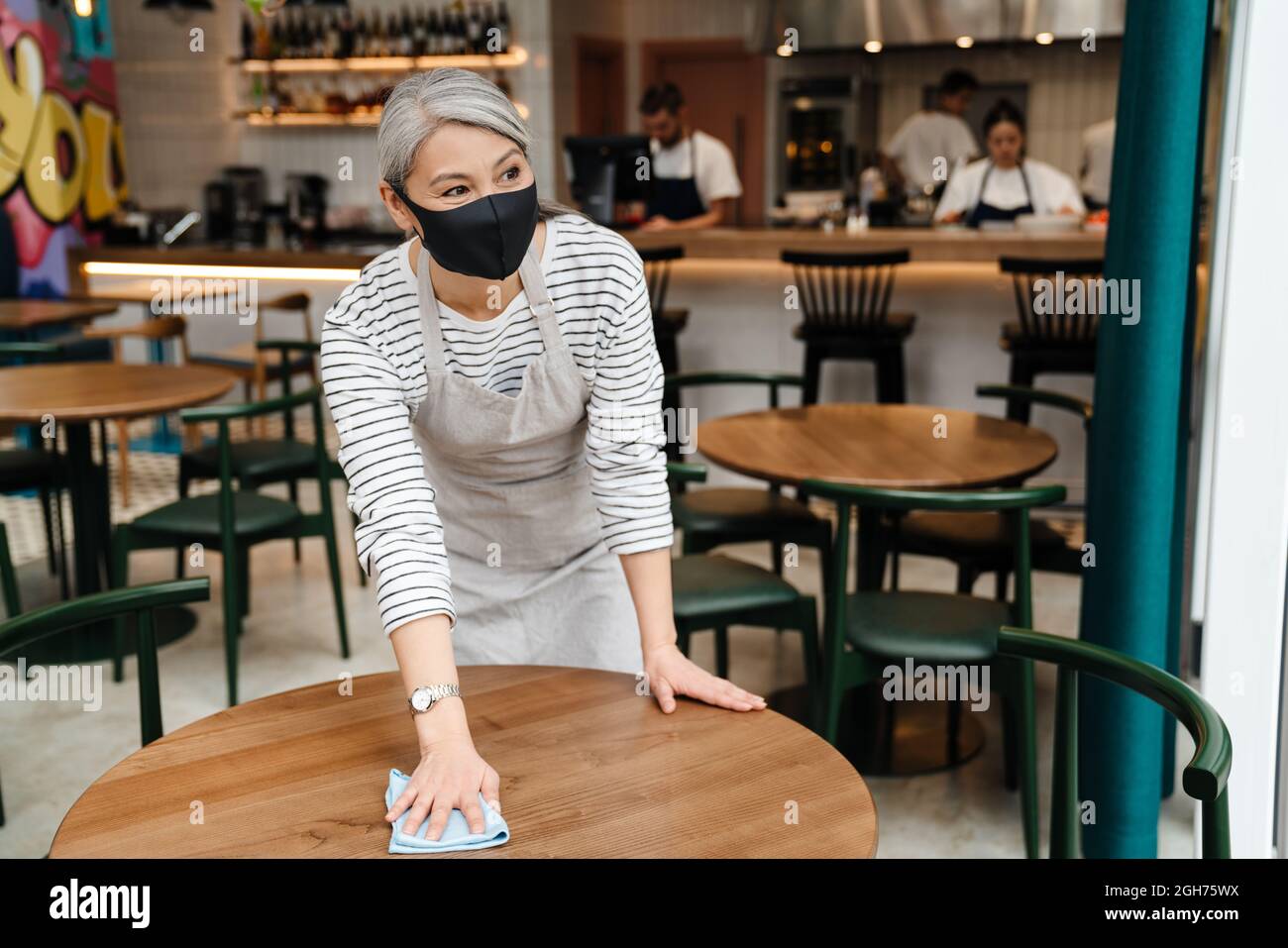 Mature waitress woman wearing face mask cleaning table in cafe indoors ...