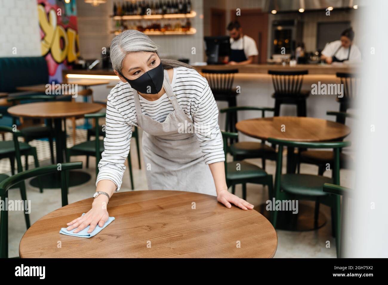 Mature waitress woman wearing face mask cleaning table in cafe indoors ...