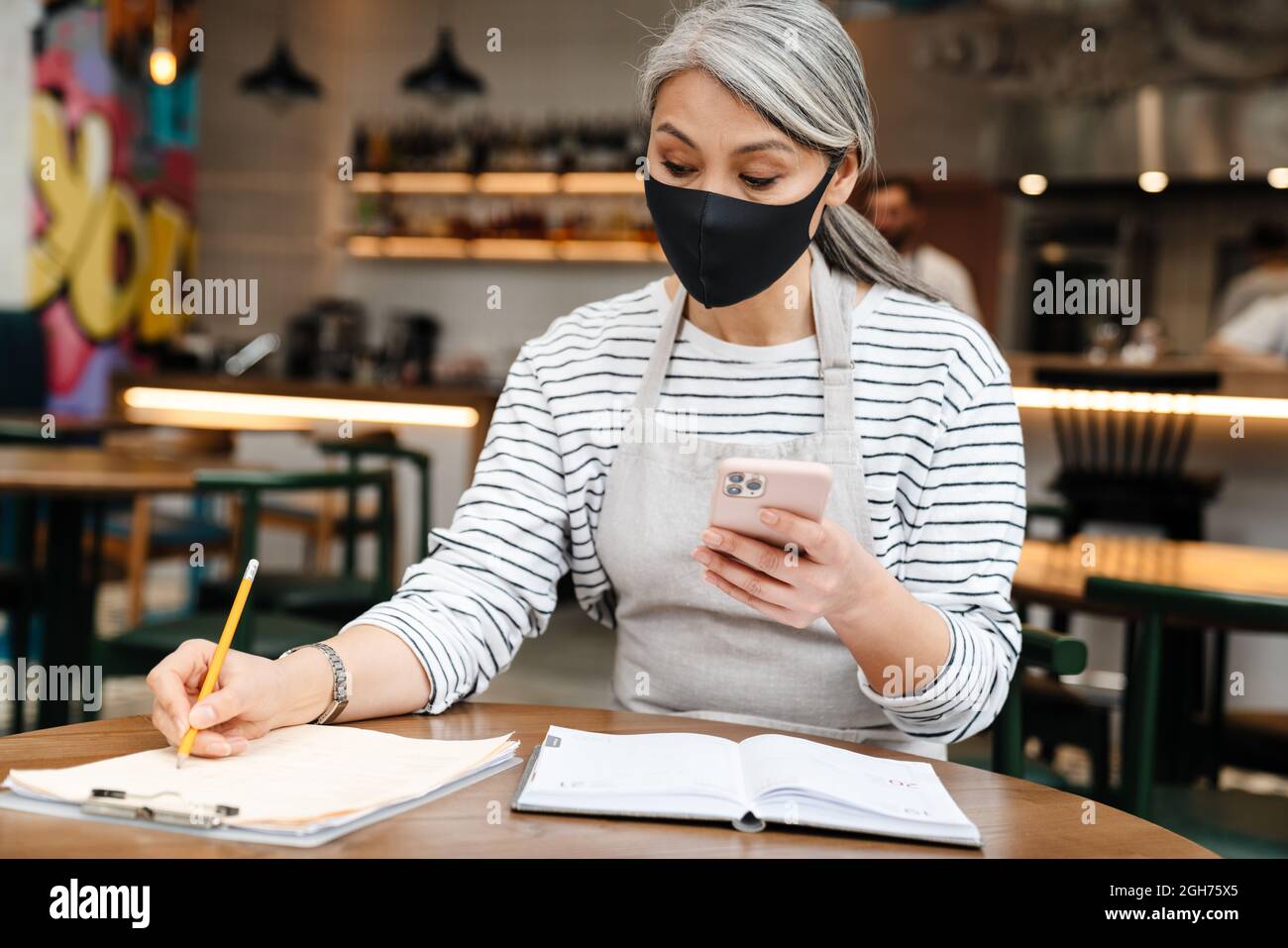 Mature waitress woman wearing face mask using cellphone while working ...