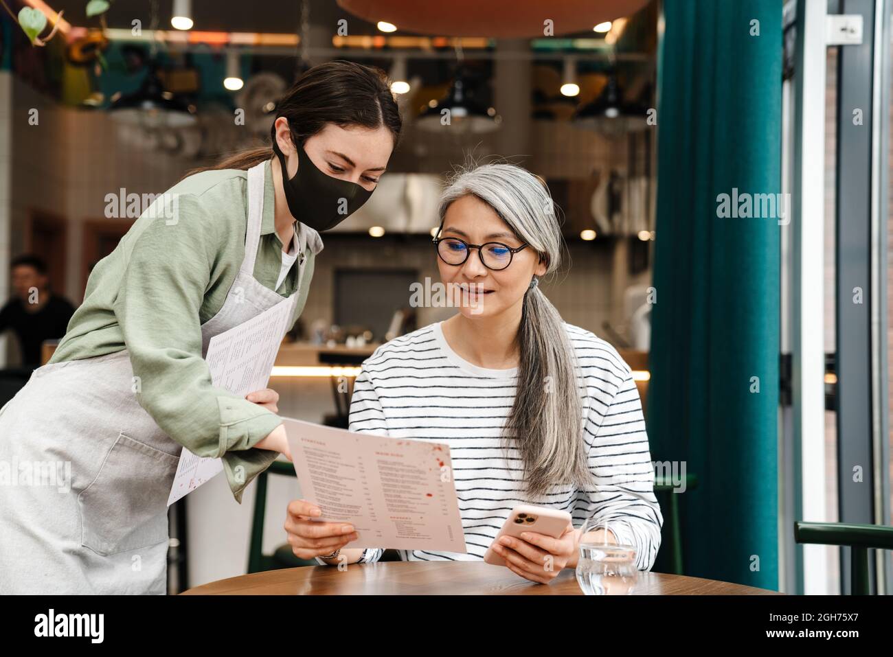 Young waitress woman wearing face mask showing menu to her client in ...