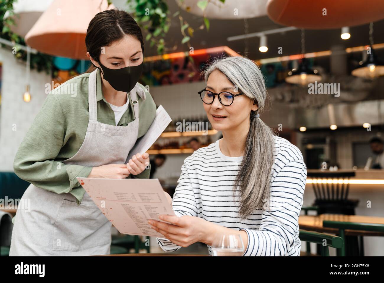 Young waitress woman wearing face mask showing menu to her client in ...