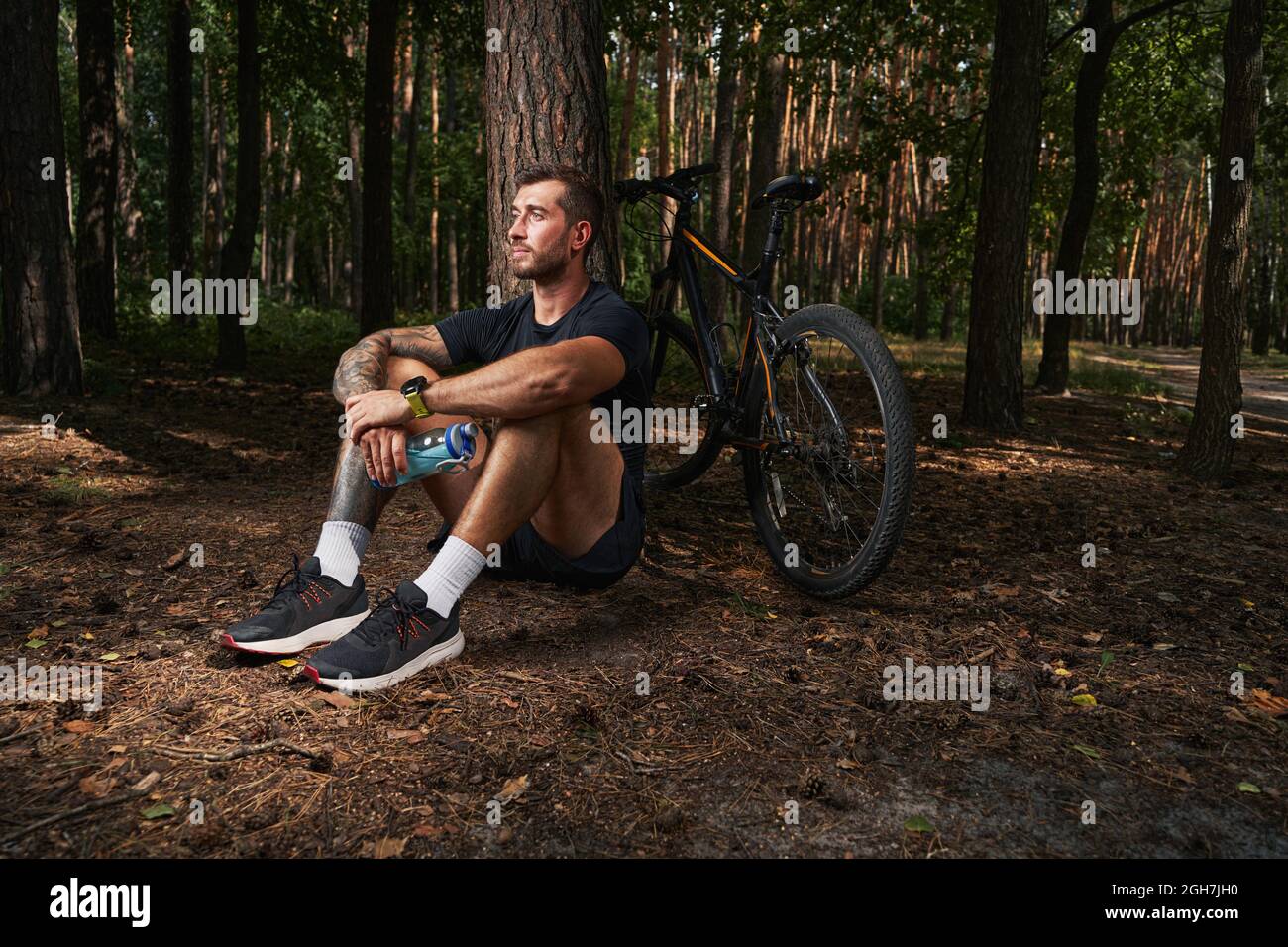 Joyful young male is relaxing under tree and drinking water after ...