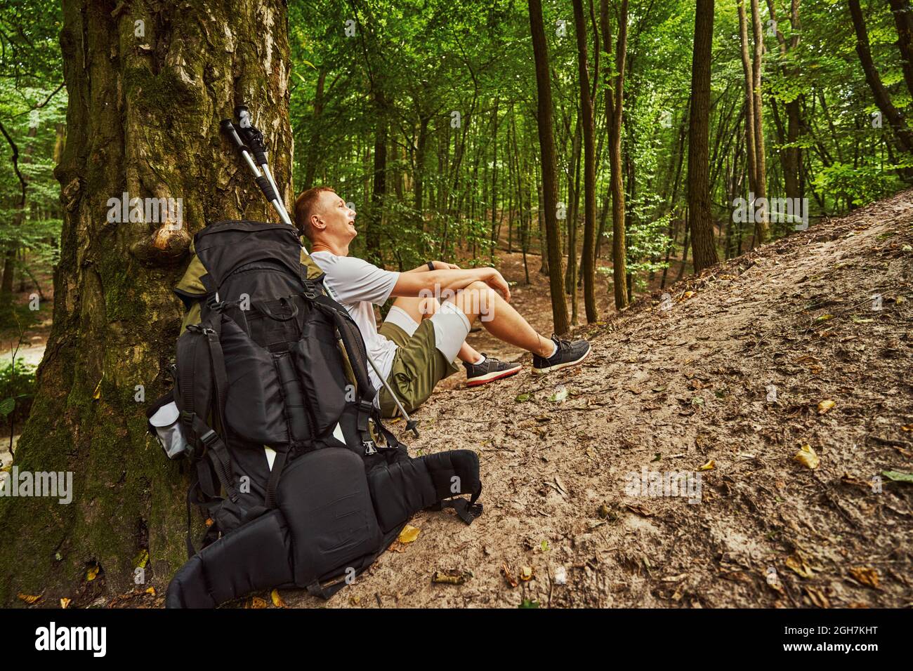 Calm active male is sitting down at tree for having rest during hiking ...
