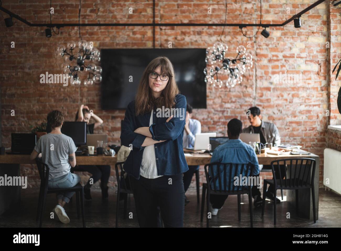 Female entrepreneur standing with arms crossed while colleagues working ...