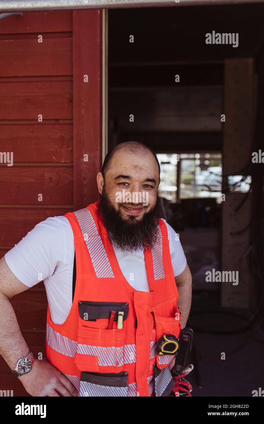 Smiling bearded male construction worker standing with hand on hip at ...