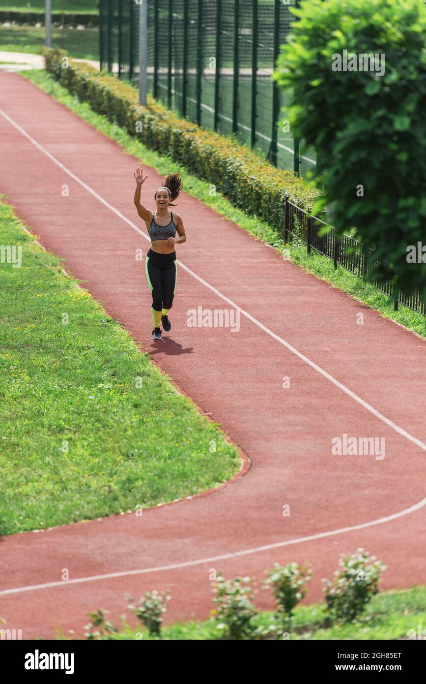 full length of happy sportswoman in wireless earphones waving hand ...