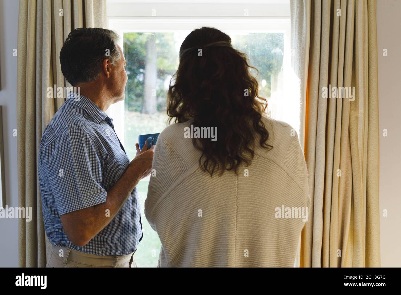 Happy senior caucasian couple standing next to window, holding cups ...