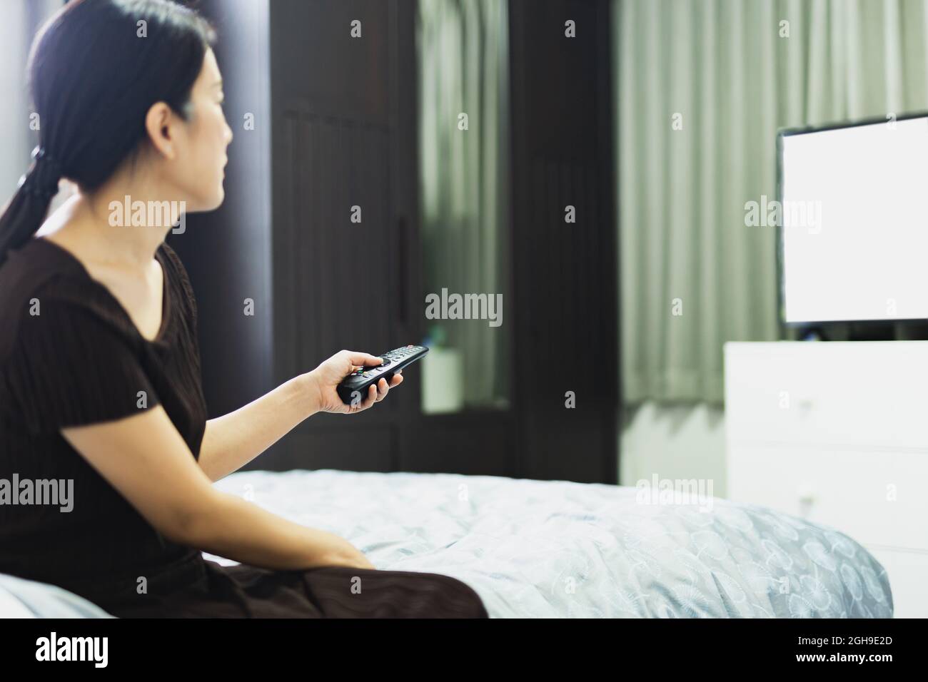 Woman watching television with remote sitting on the bed at home Side ...