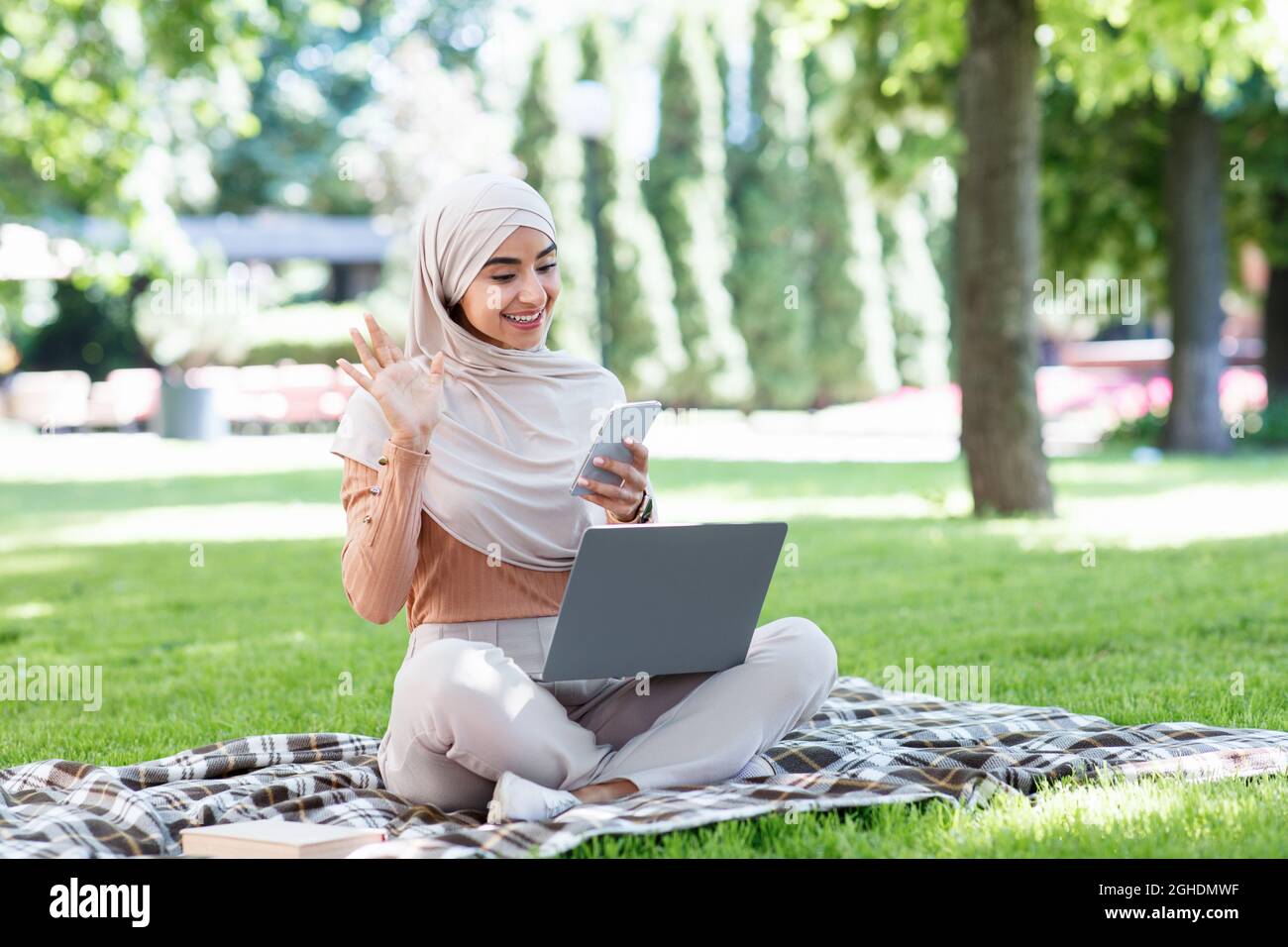 Smiling millennial arab woman in hijab with laptop, calls on smartphone ...