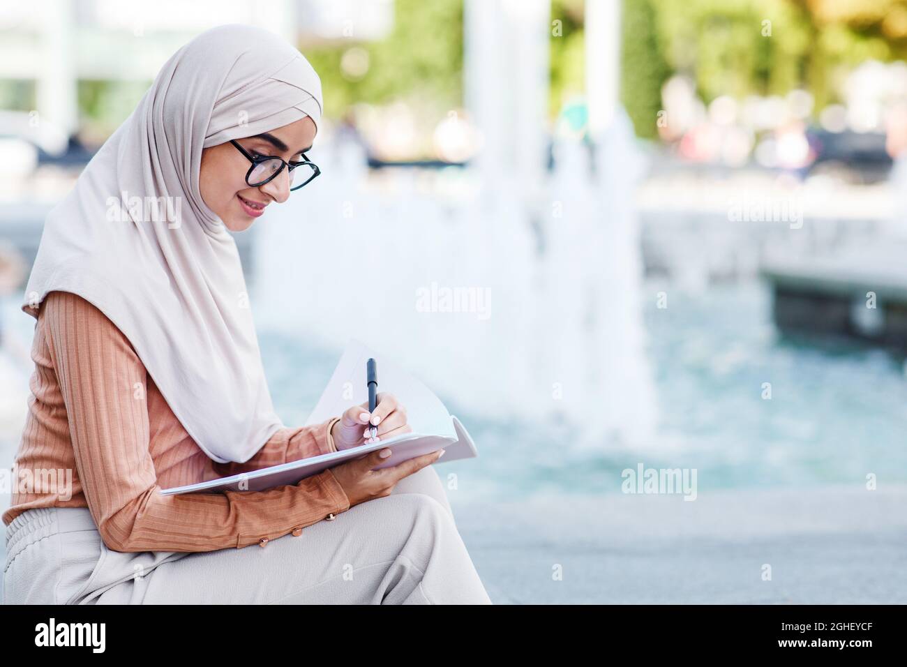 Cheerful cute young middle eastern female in hijab writing in notebook ...