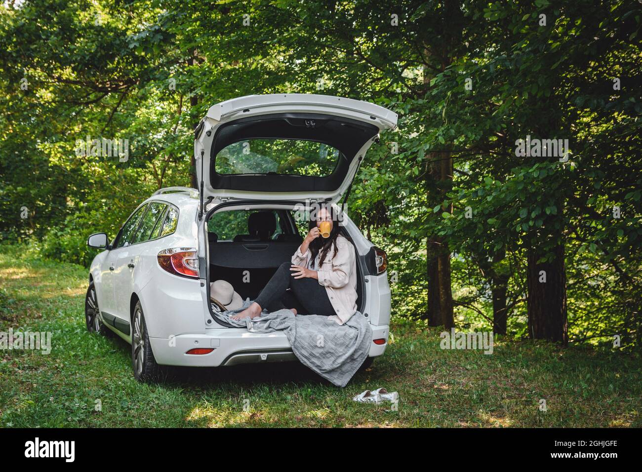 Happy young woman on a camping road trip sitting in car boot drinking ...