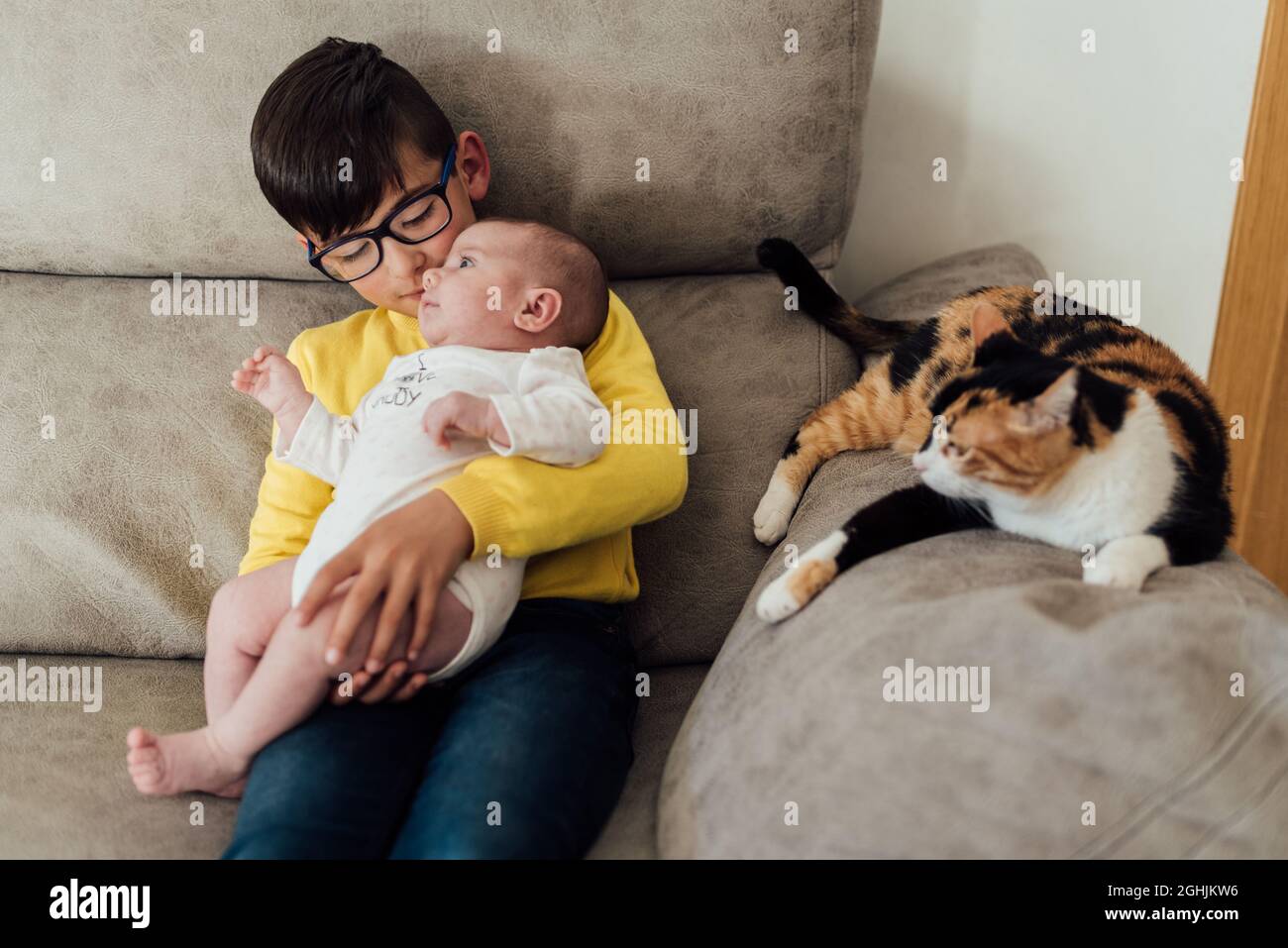 Boy holding his little baby sister in his arms while sitting on a sofa ...