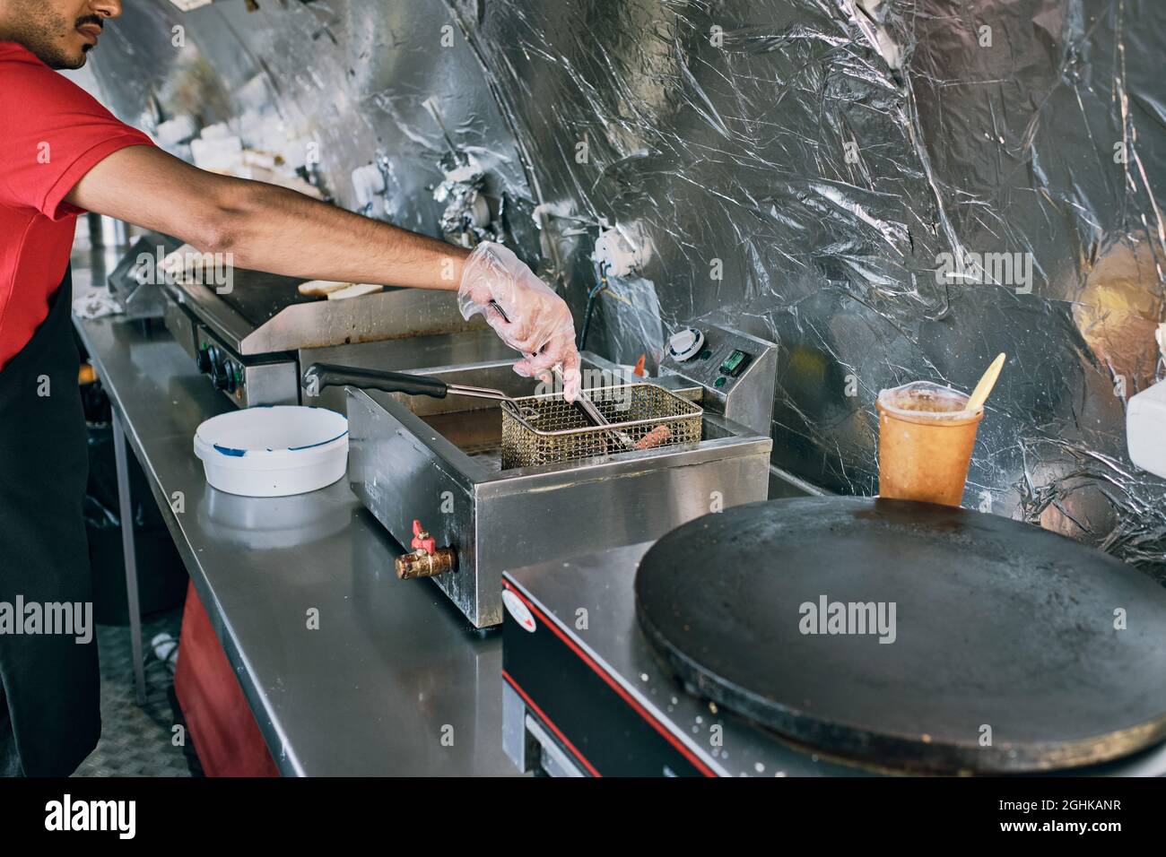 Gloved young man putting sausage into frying equipment while cooking ...