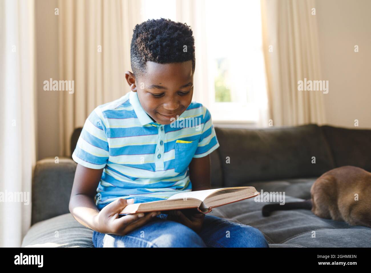 Smiling african american boy reading book and sitting on couch with cat ...