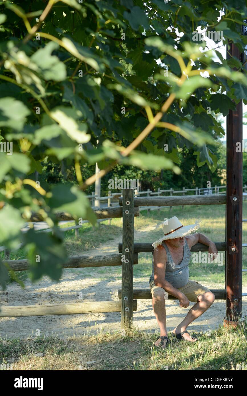 Vertical shot of a villager sitting on the wooden fence of a round pen ...