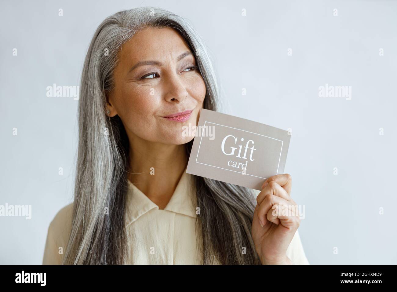 Positive silver haired Asian woman holds gift card standing on light ...