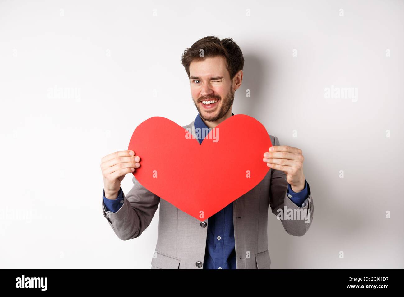 Charismatic young man winking and smiling, showing big red heart cutout ...