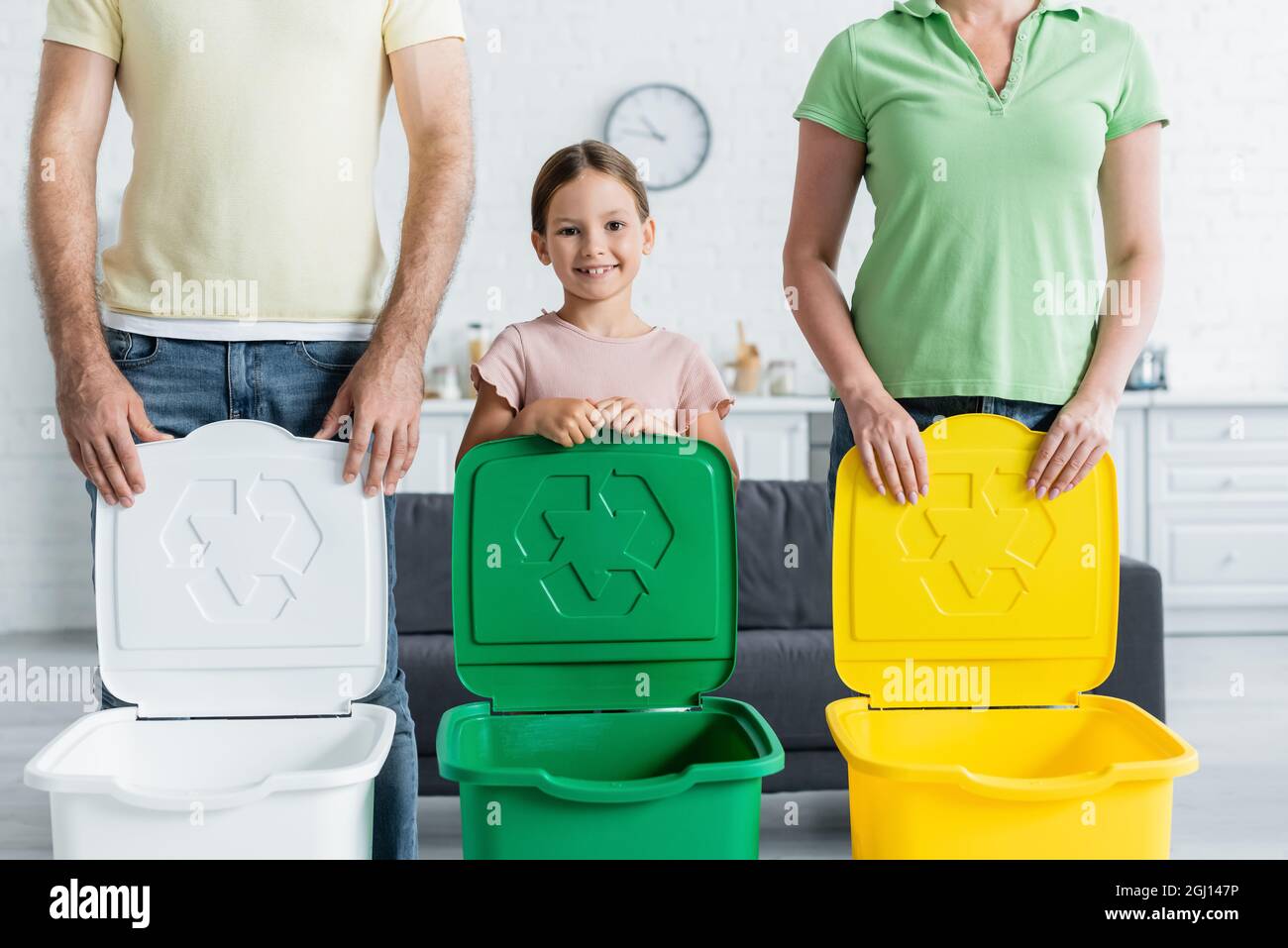 Smiling girl looking at camera near parents and trash bins with recycle ...