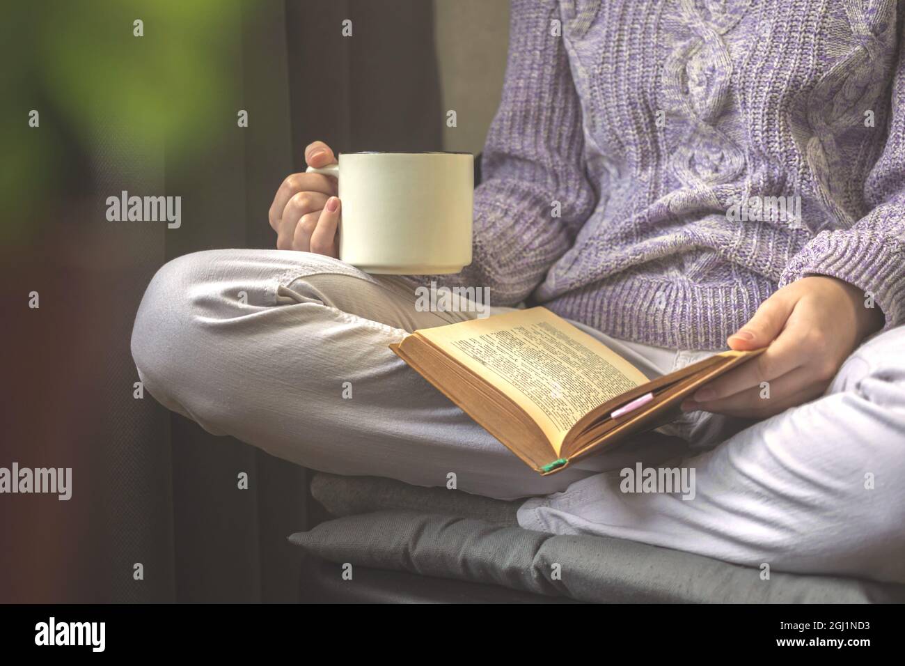 Young woman reading book with tea cup in hand sitting on chair at home ...
