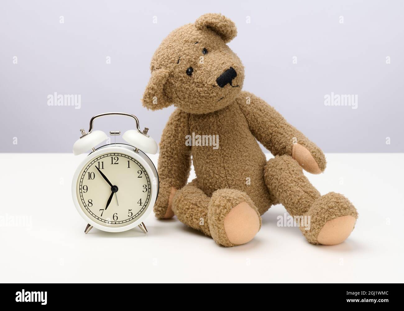 brown teddy bear sits on a white table and a round alarm clock, at five ...
