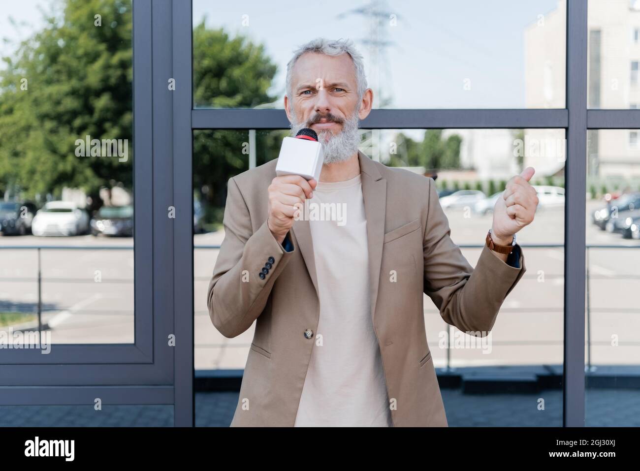 bearded reporter in blazer holding microphone and pointing with thumb ...