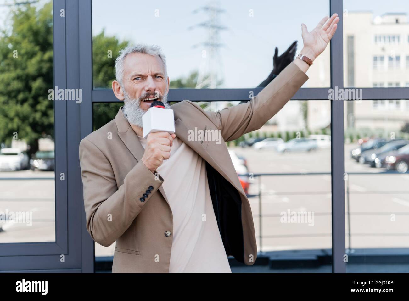 bearded reporter in blazer holding microphone and pointing with hand at ...