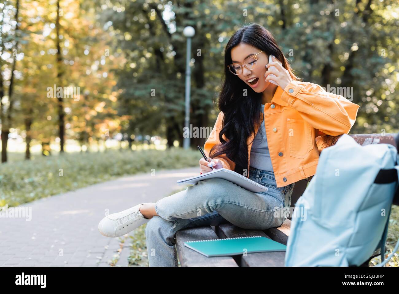 astonished asian woman writing in notebook while talking on smartphone ...