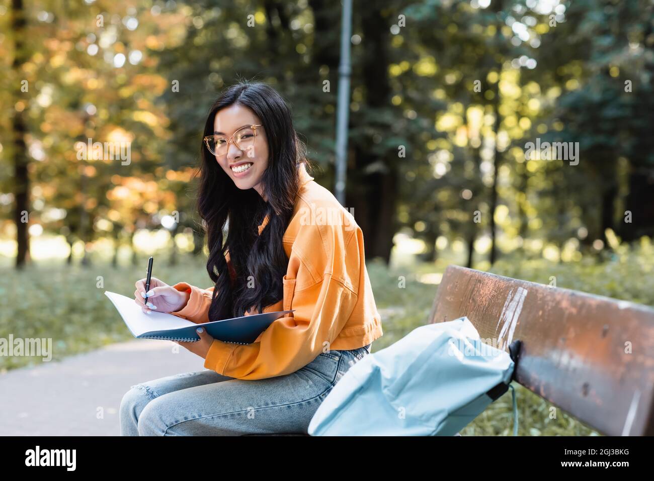 happy asian woman writing in notebook and smiling at camera on bench in ...