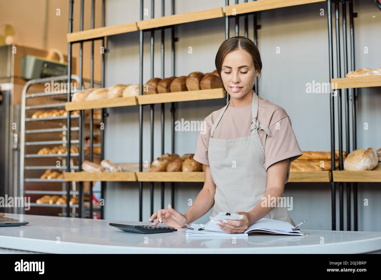 Content young Caucasian female clerk in apron standing at counter in ...