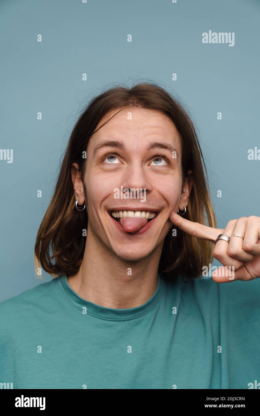 Young White Man Showing His Tongue And Pointing Finger At His Face Isolated Over Blue Wall Stock