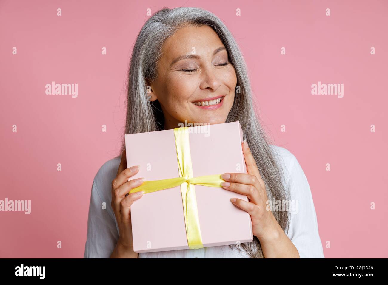 Cheerful mature Asian lady holds gift box with ribbon posing on pink ...