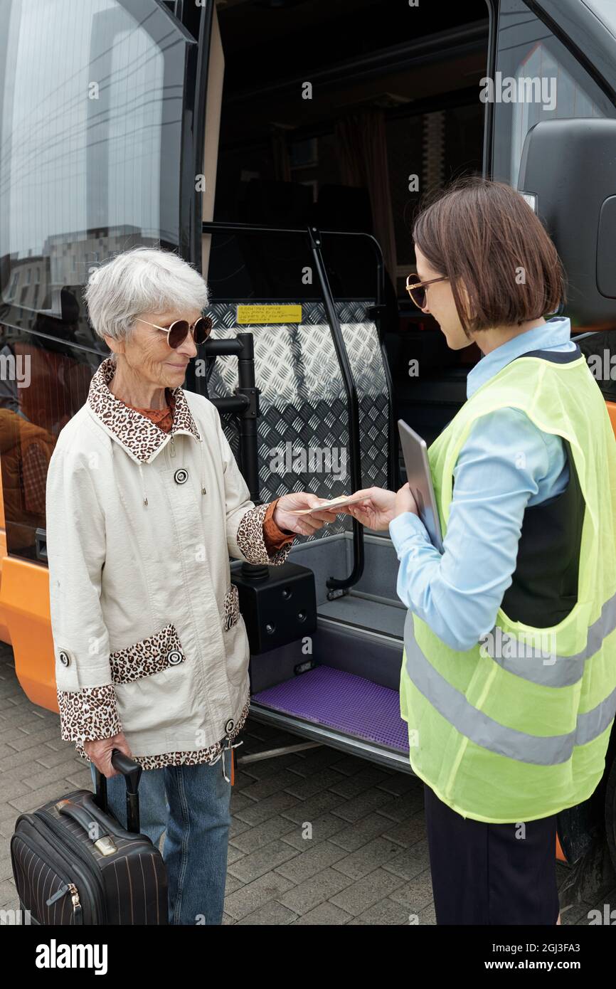 Smiling senior Caucasian woman with suitcase giving ticket to bus ...