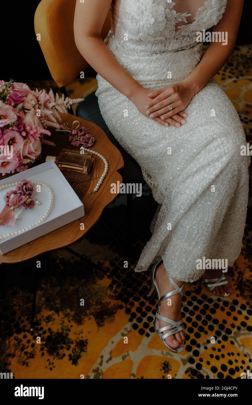 Bride sitting on chair next to a table with bouquet of flowers, perfume ...
