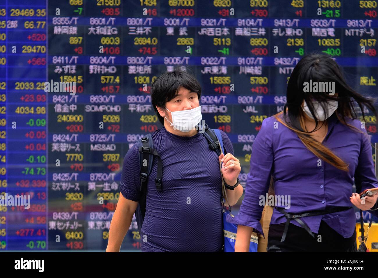 People wearing masks walk away from an ic board showing stock market ...
